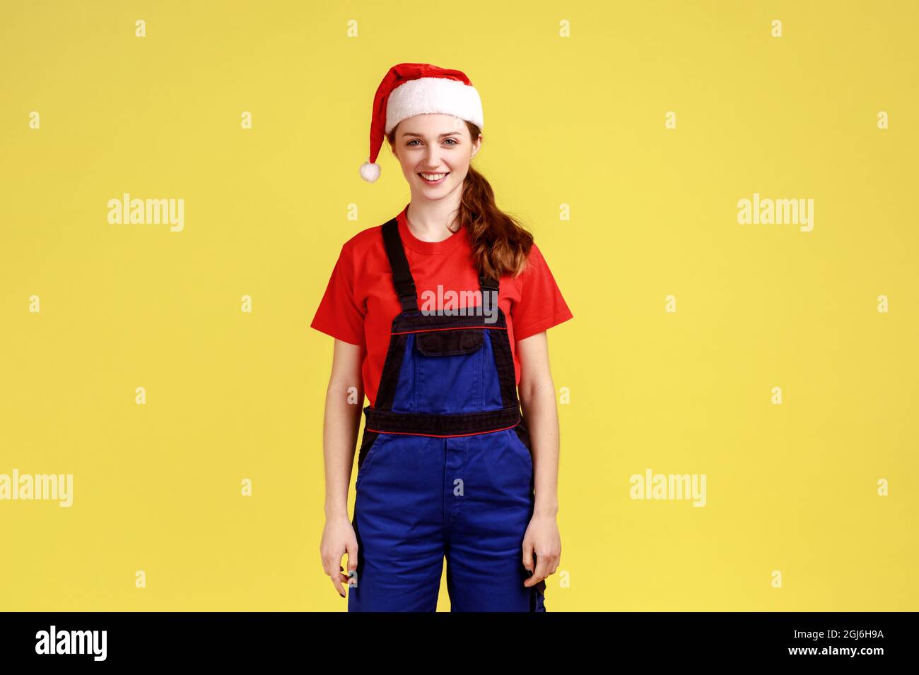 Happy handy woman in blue overalls and santa hat standing looking at ...