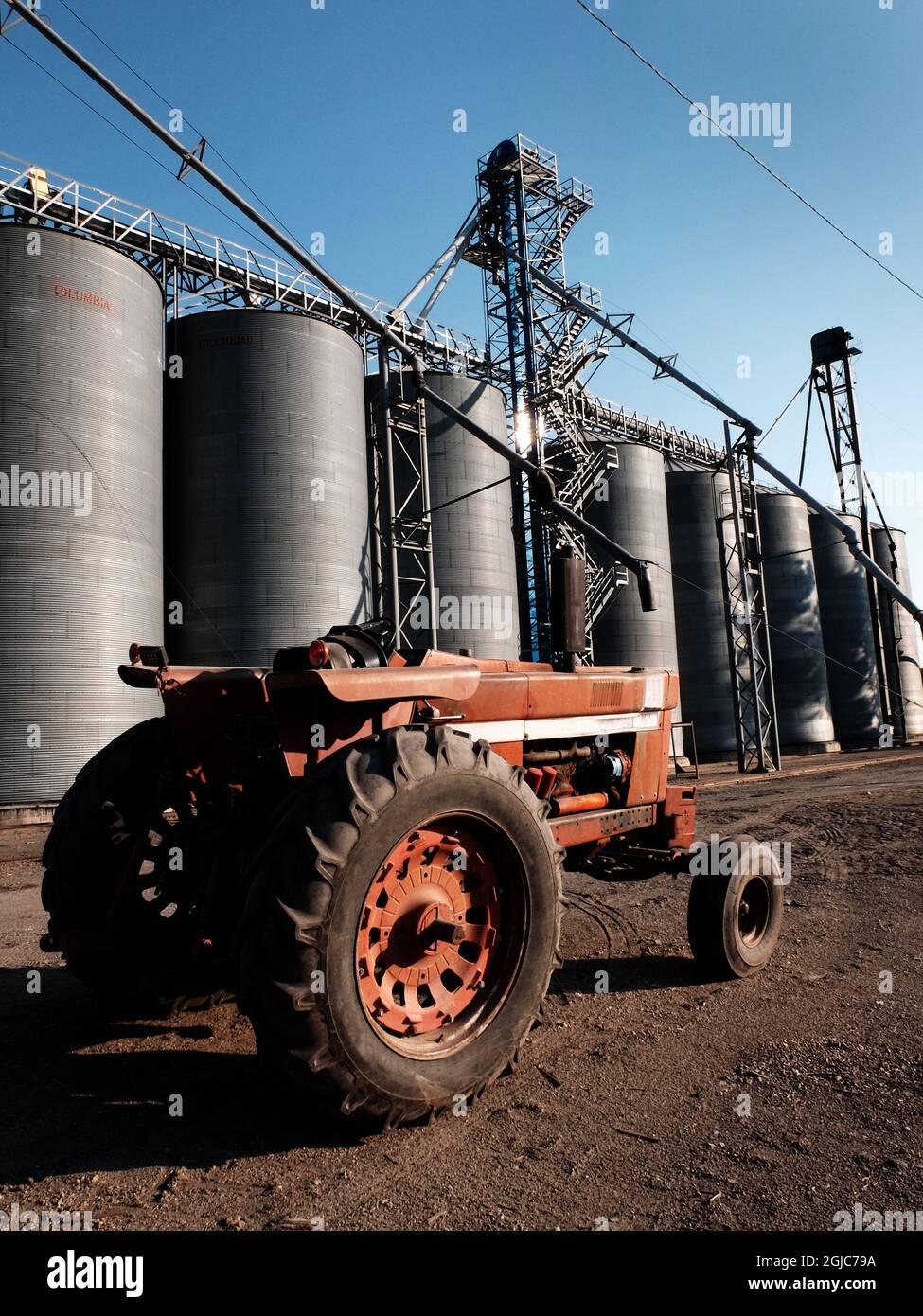 Old red work tractor parked in front of several large steel grain silos ...