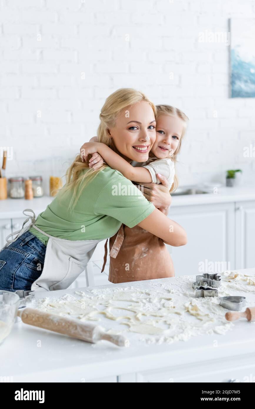 happy mom and daughter in aprons looking at camera while embracing near ...