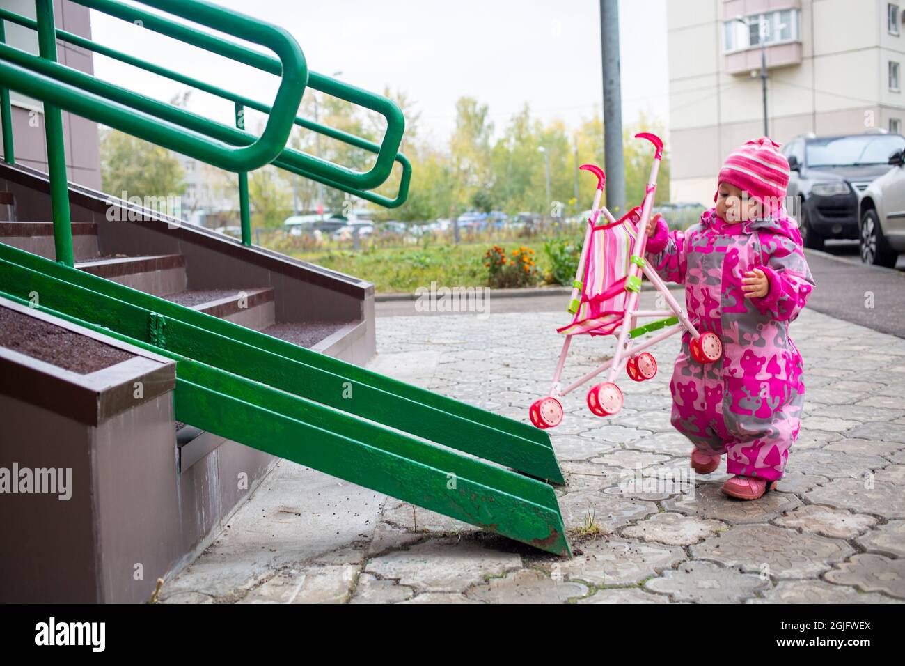 cute toddler drags a toy stroller along the ramp of the stairs. Slope ...