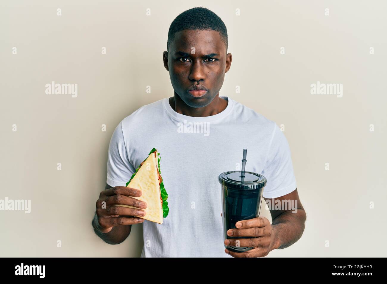Young african american man eating sandwich and drinking soda skeptic ...