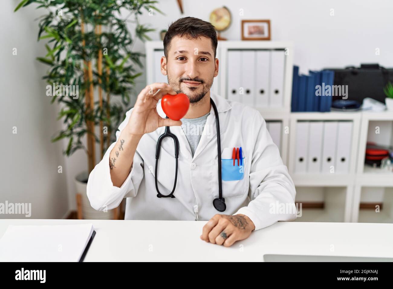 Young doctor holding heart at medical clinic looking positive and happy ...