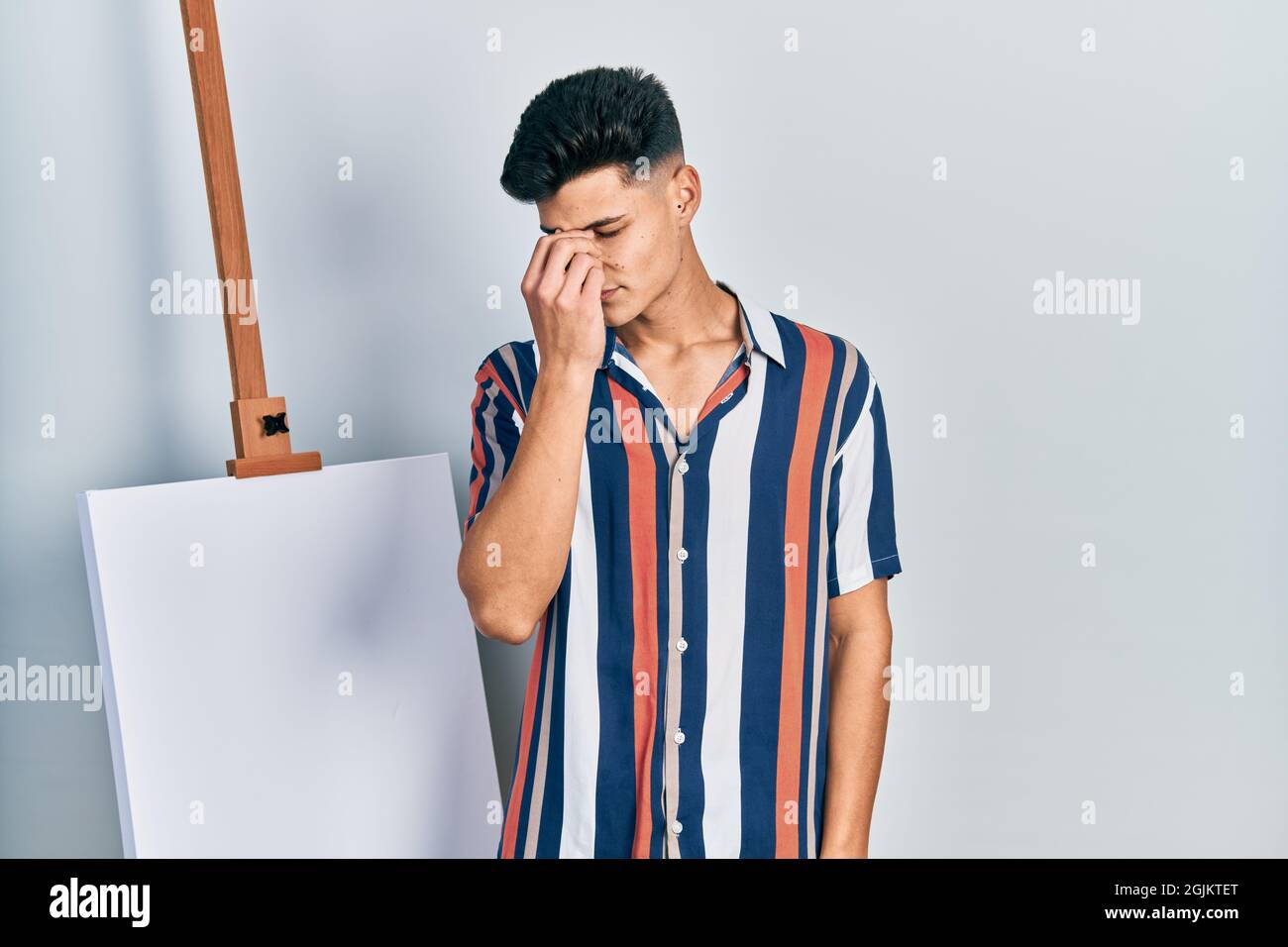 Young hispanic man standing close to empty canvas tired rubbing nose ...