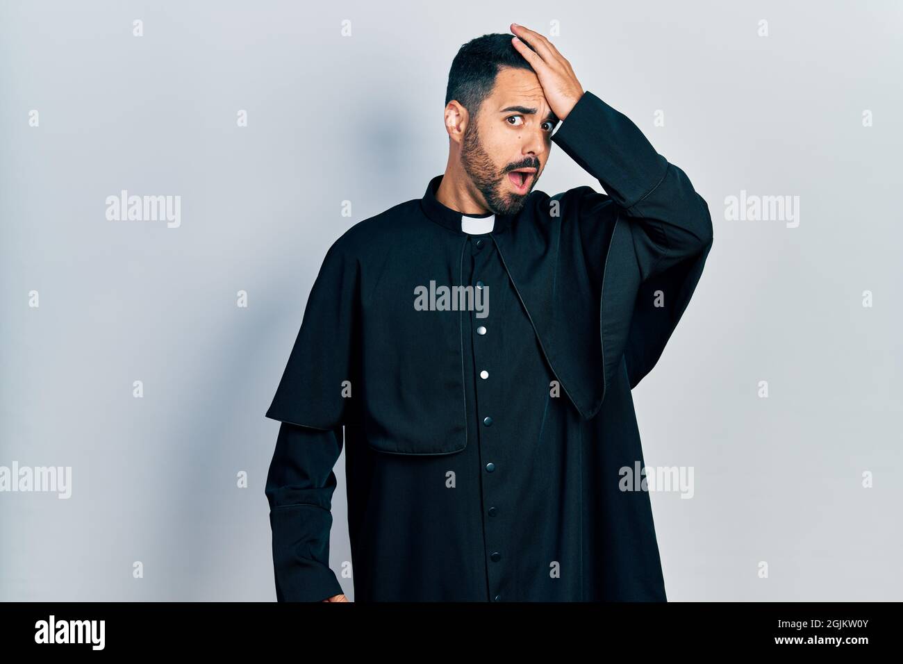 Handsome hispanic man with beard wearing catholic priest robe surprised ...