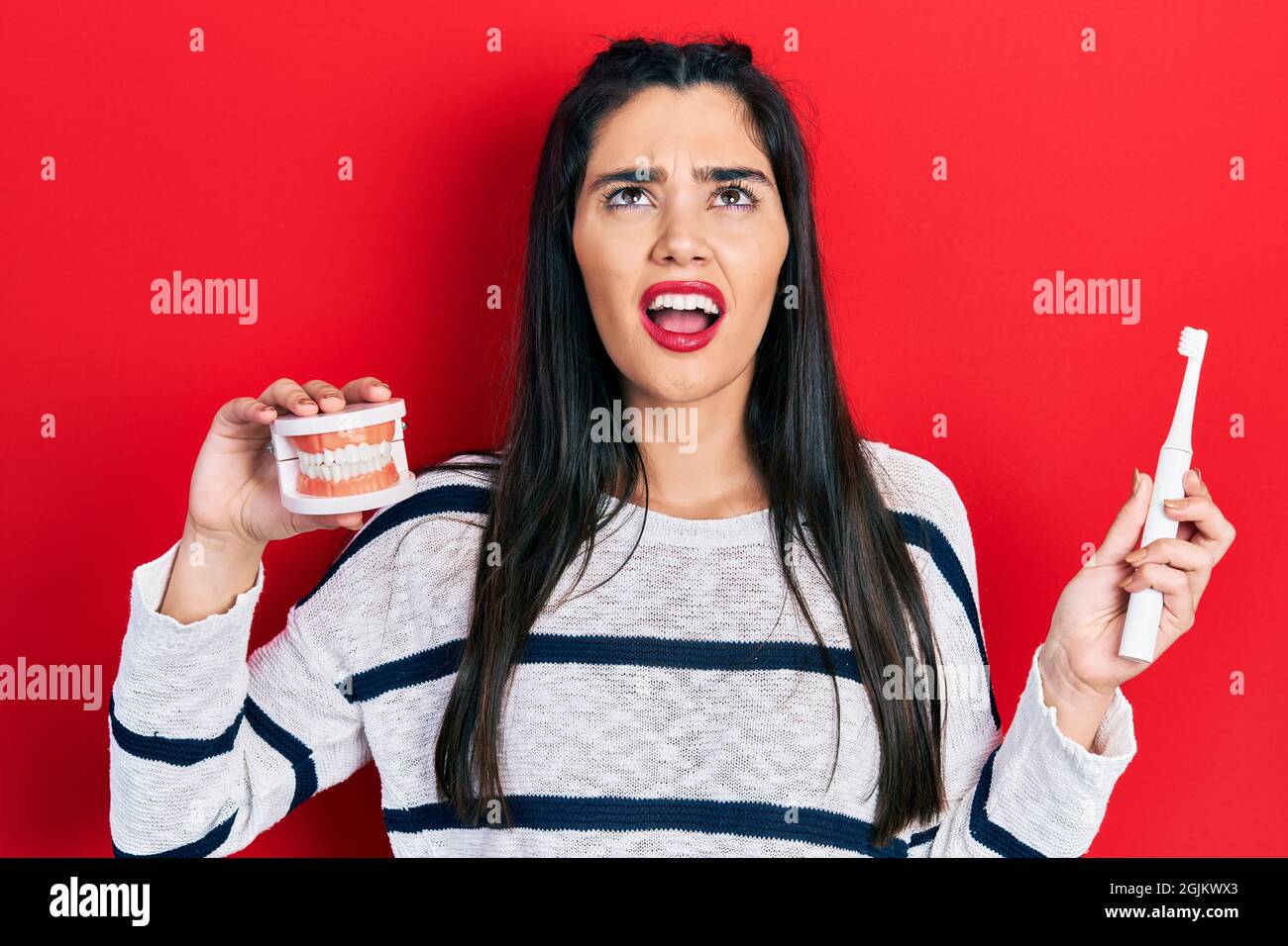 Young hispanic girl holding electric toothbrush and denture angry and mad screaming frustrated ...