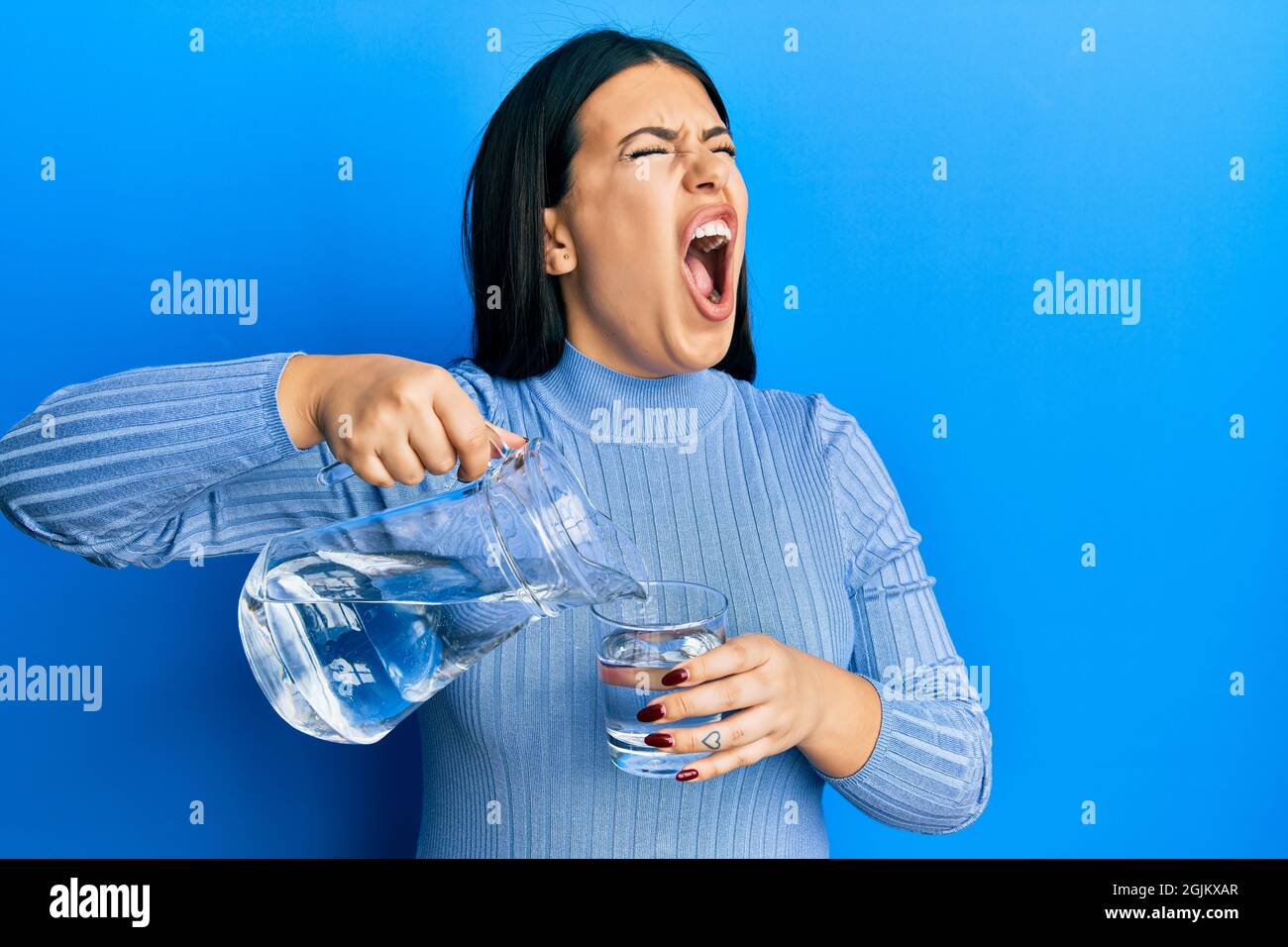 Beautiful brunette woman pouring water in glass angry and mad screaming ...