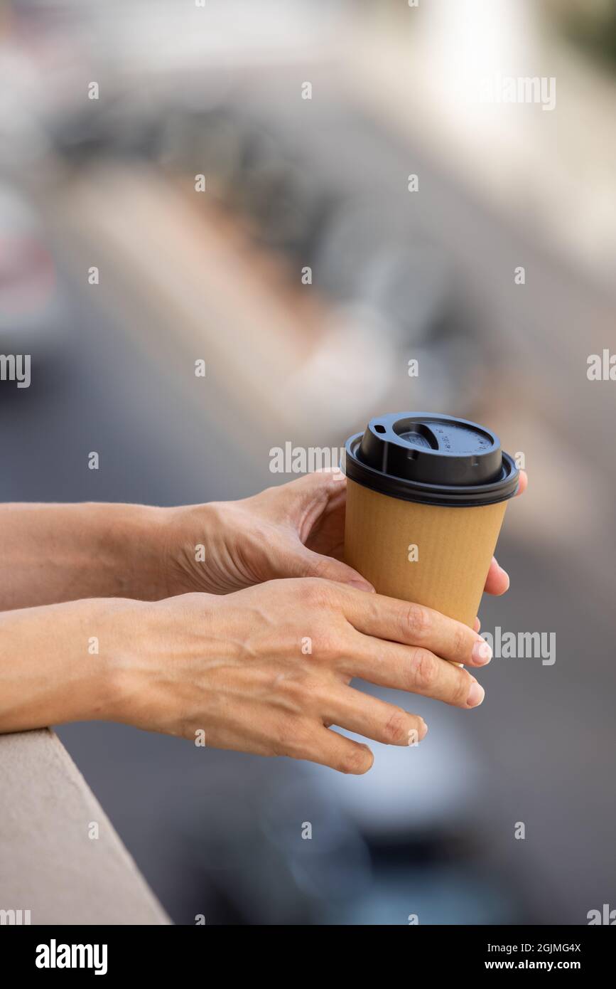 Hands holding a single use paper coffee cup. Young woman in a balcony ...