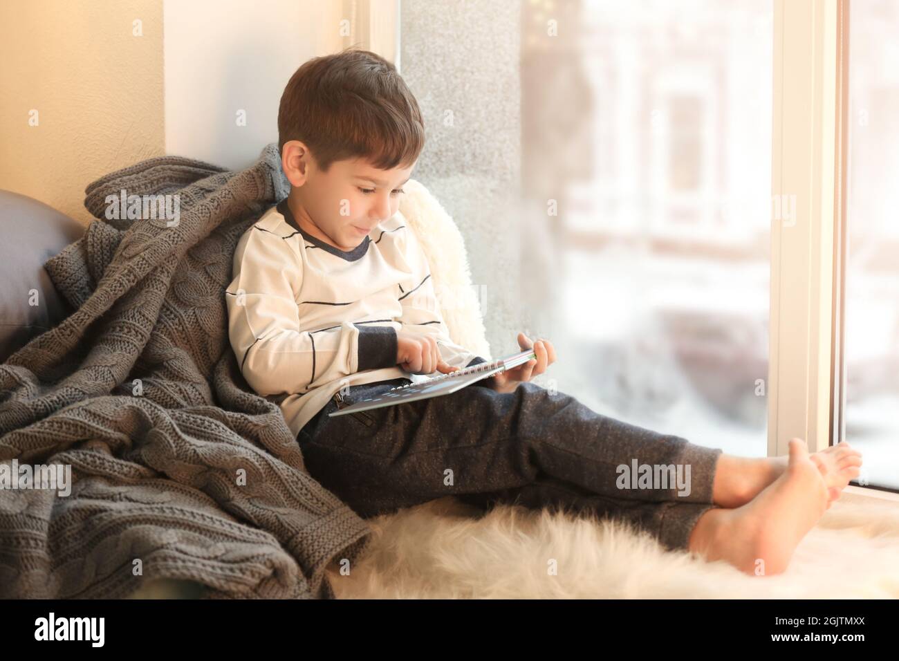 Cute little boy reading book while sitting on window sill at home Stock ...