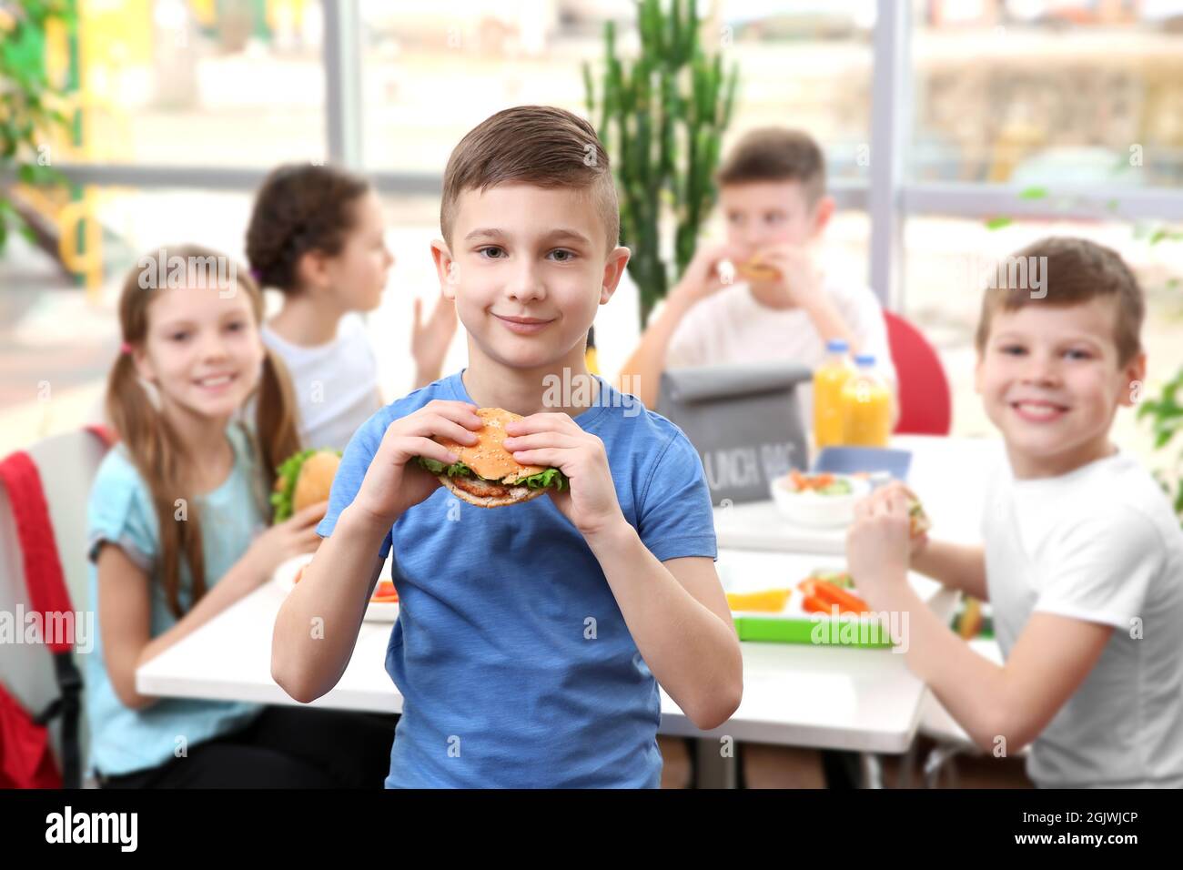 Cute boy eating hamburger and children sitting at table in school ...