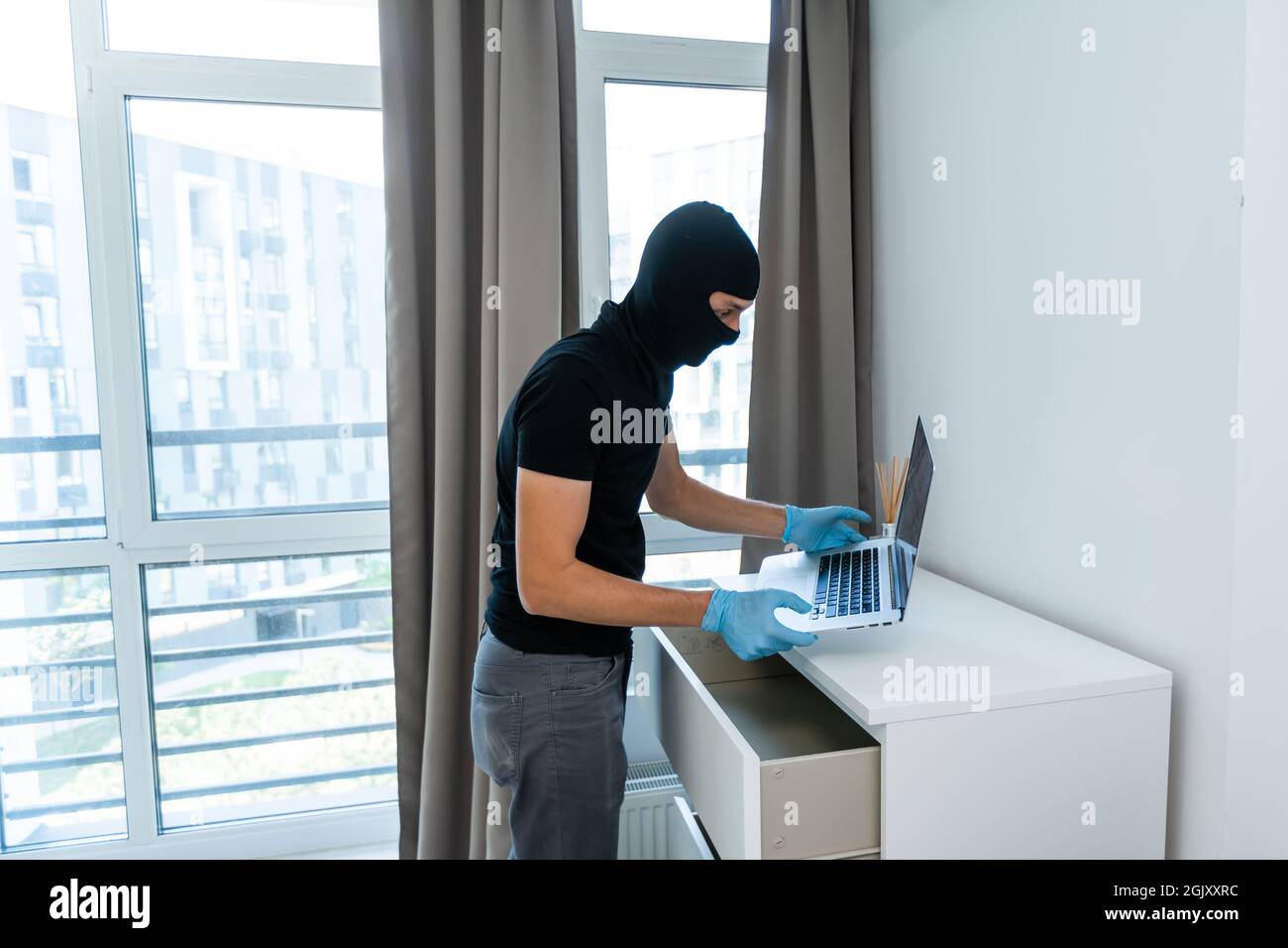 Thief with black balaclava stealing laptop. The burglar commits a crime ...