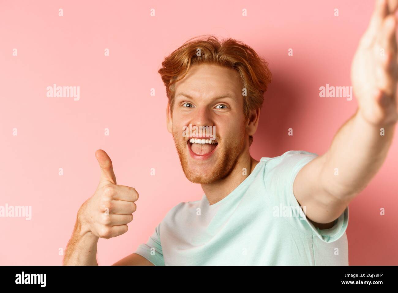 Excited redhead man tourist taking selfie and showing thumbs-up, holding camera with stretched ...