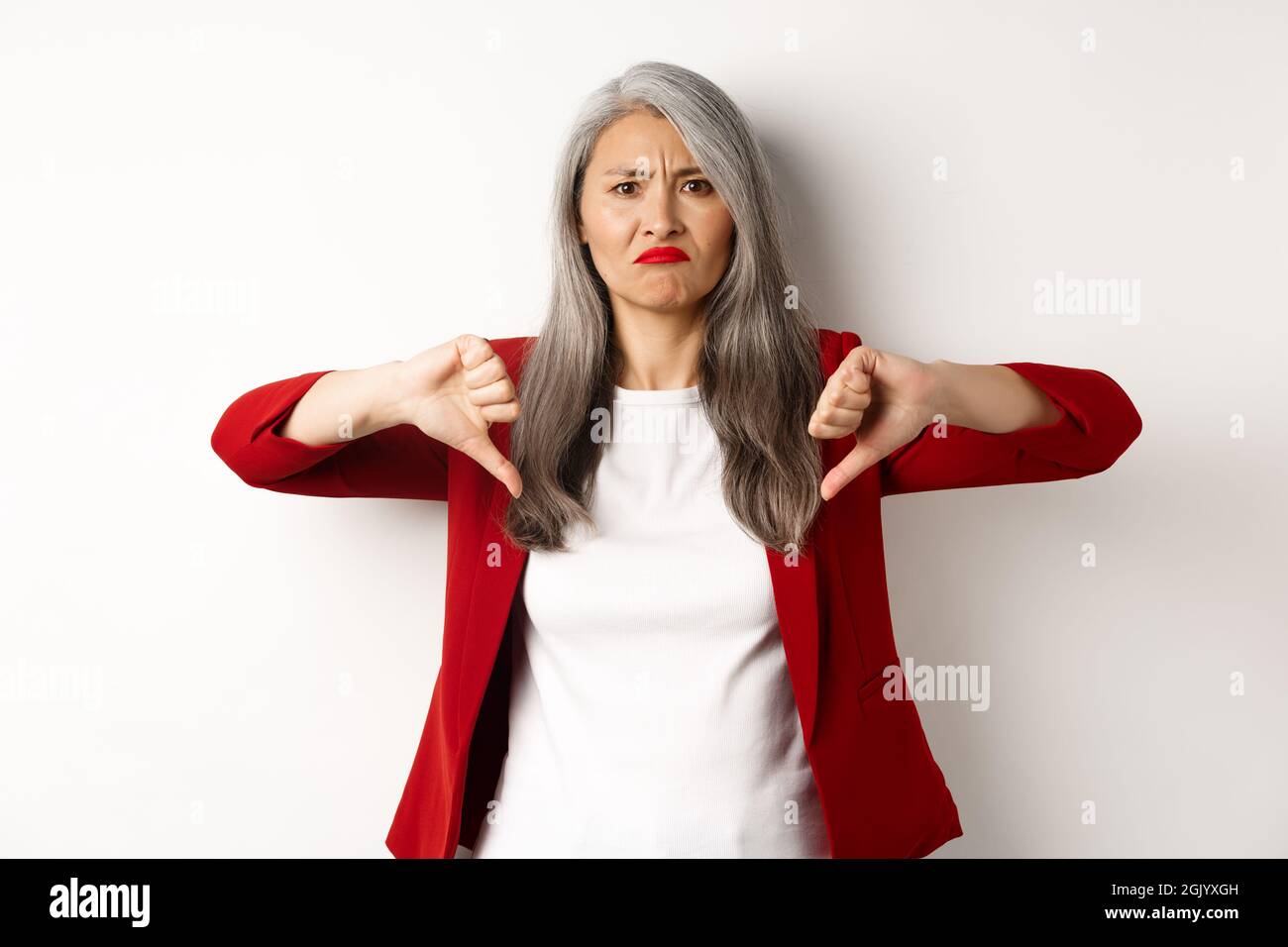 Disappointed elderly woman in red blazer showing thumbs-down, grimacing ...
