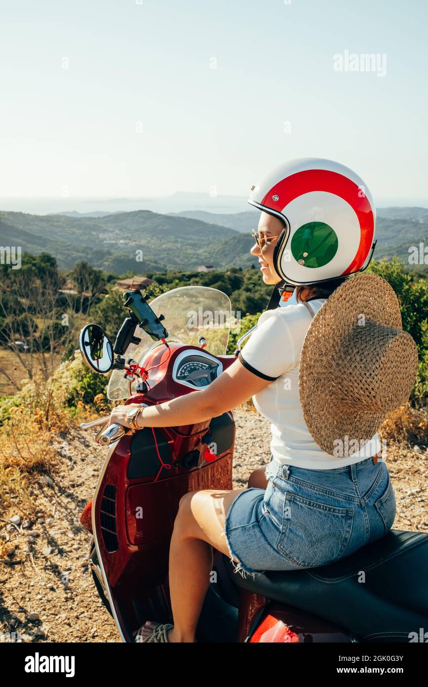 Woman sitting on retro scooter or motor bike in red color, traveling in mountains on island ...