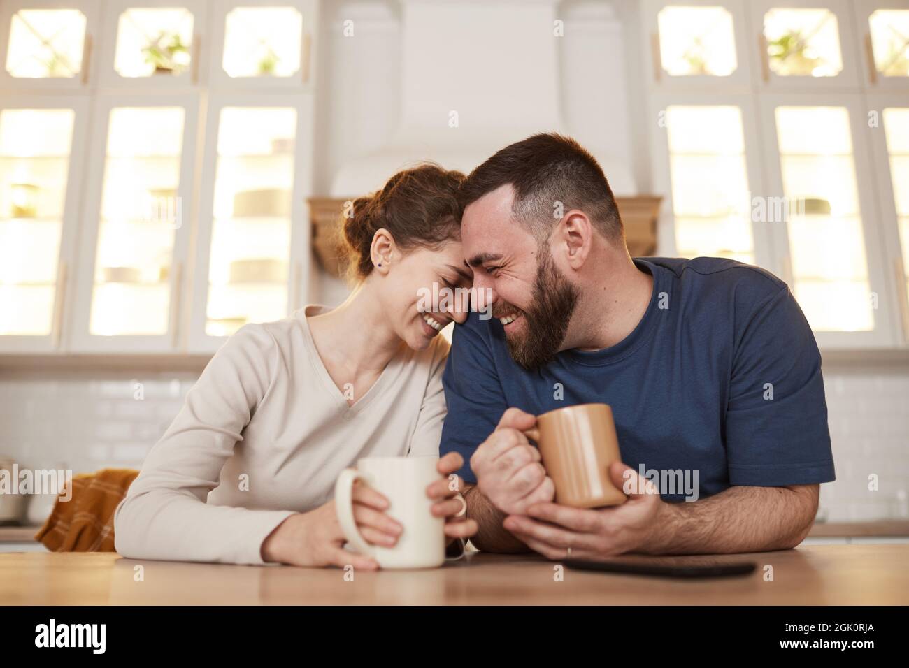 Happy affectionate young Caucasian couple leaning on kitchen table and ...