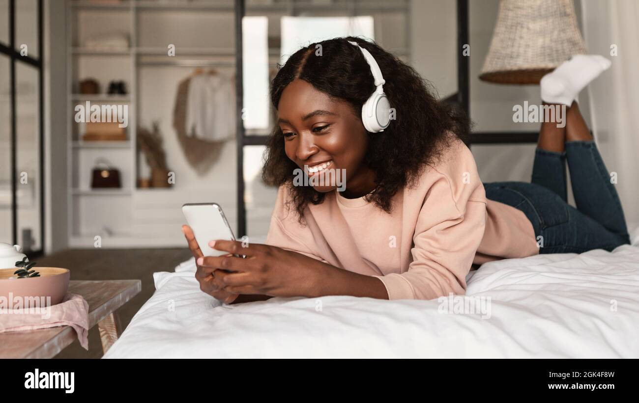 Happy African Lady Using Phone Wearing Headphones Lying In Bedroom ...
