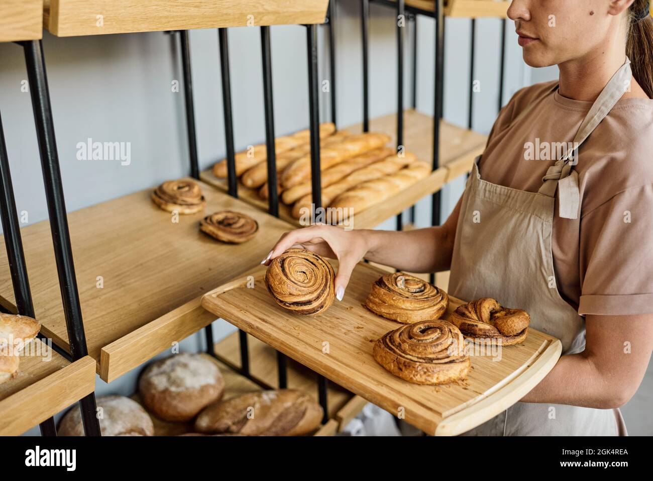 Serious young female bakery worker in apron putting pastry from tray ...