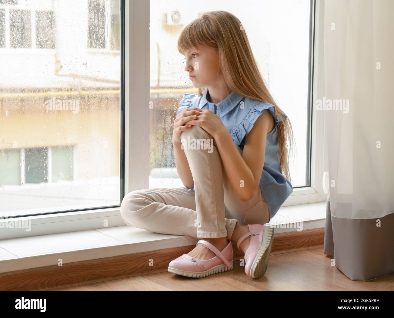Sad little girl sitting on window sill at home Stock Photo - Alamy