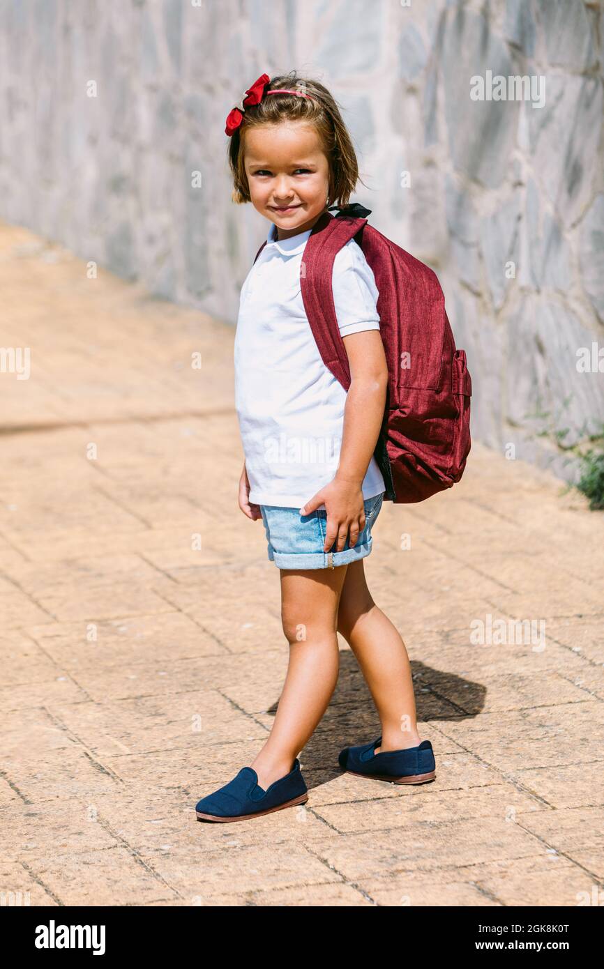 Side view of schoolchild with backpack on pavement looking at camera in ...