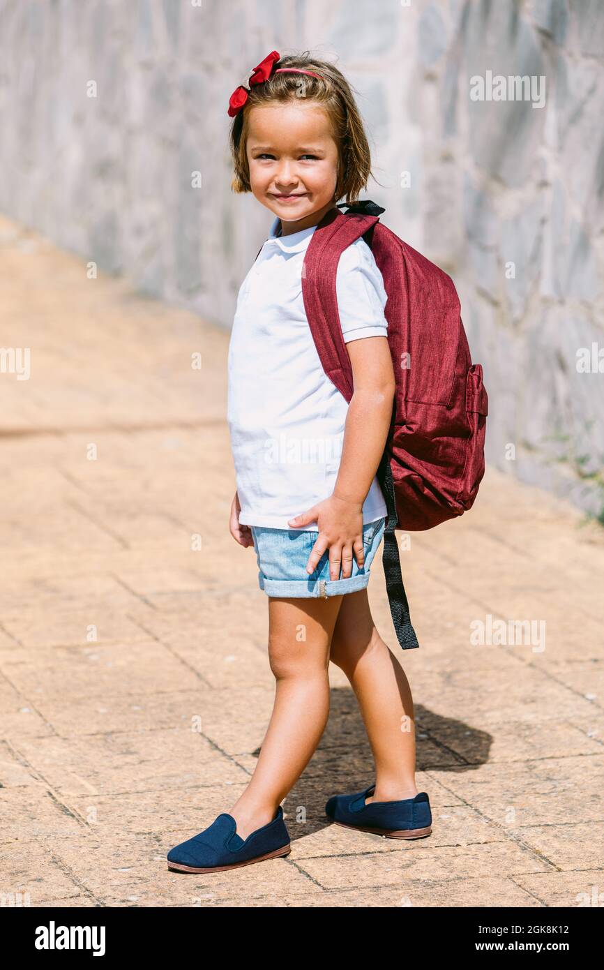 Side view of schoolchild with backpack on pavement looking at camera in ...