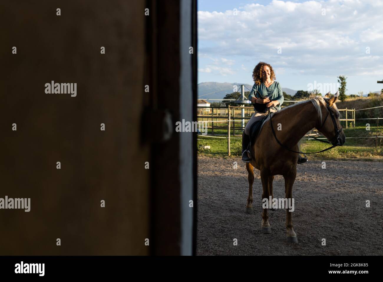 Smiling female sitting on horse in stall in countryside riding school ...