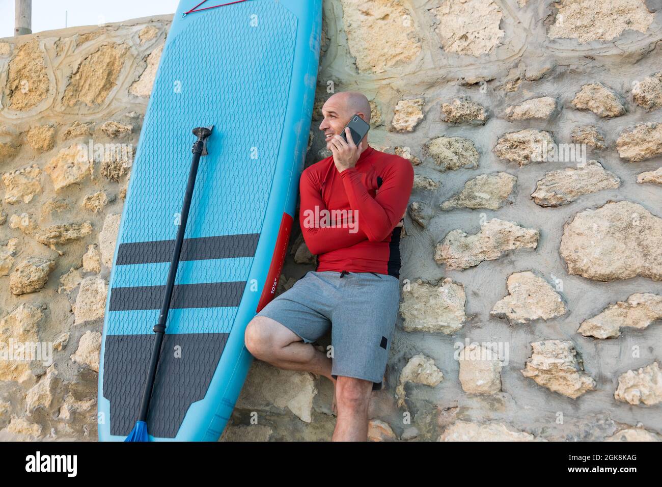 Male surfer in wetsuit leaning on stone wall speaking on smartphone ...