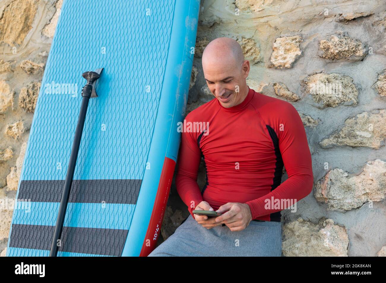 Male surfer in wetsuit leaning on stone wall browsing on smartphone ...