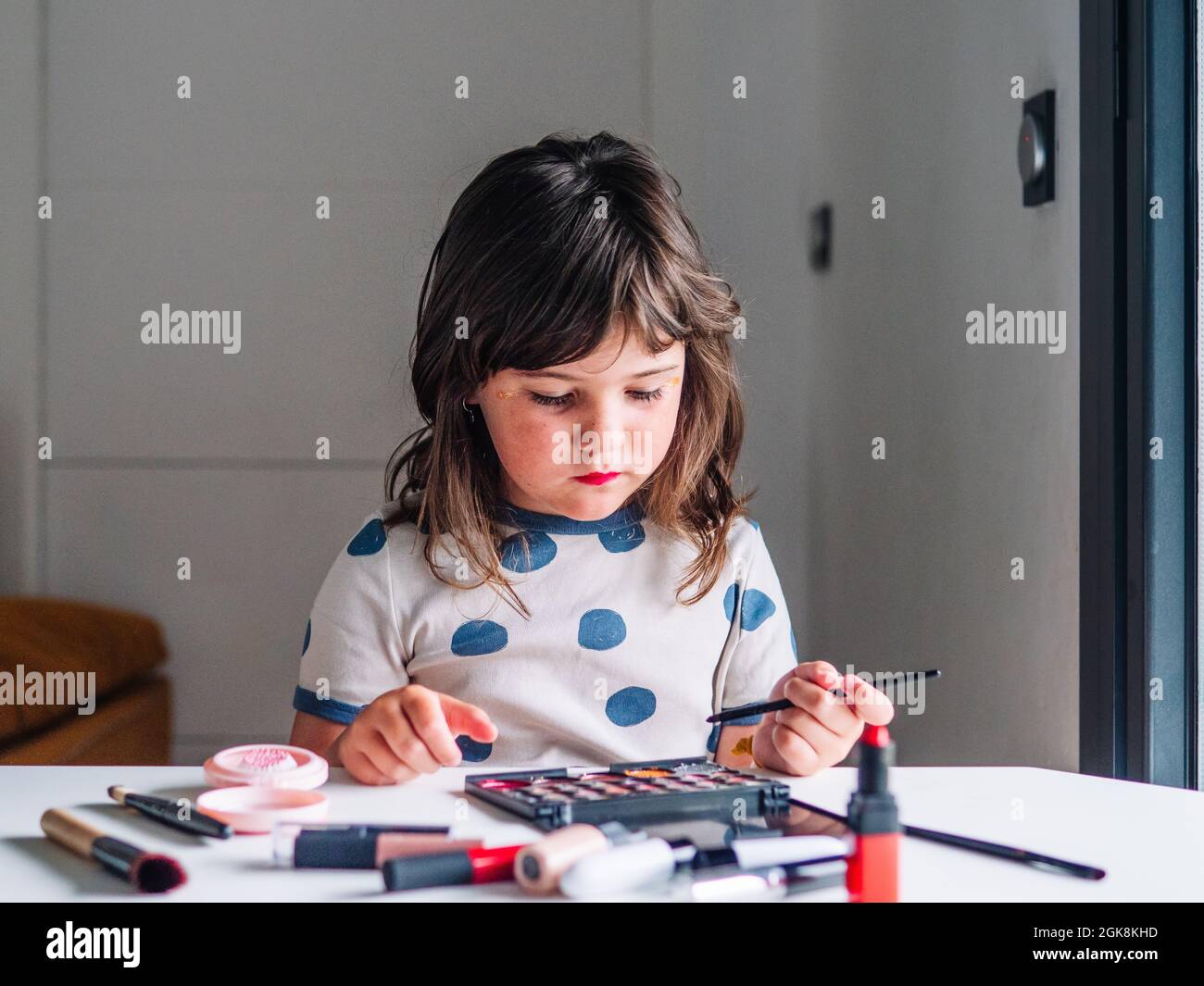 Kid with applicator making up face at table with assorted cosmetic ...