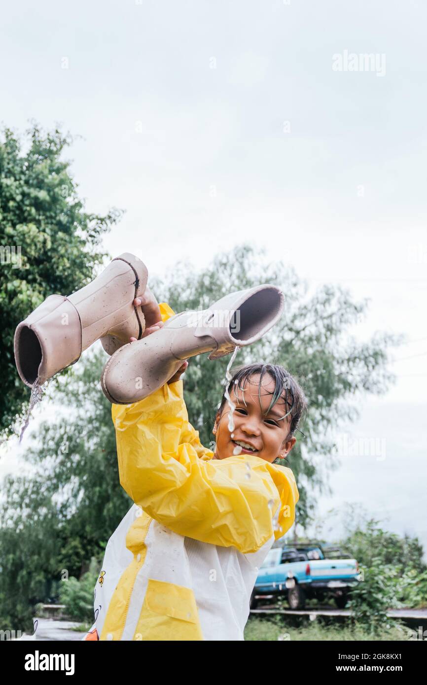 Smiling ethnic kid in slicker pouring aqua from gumboots while looking ...