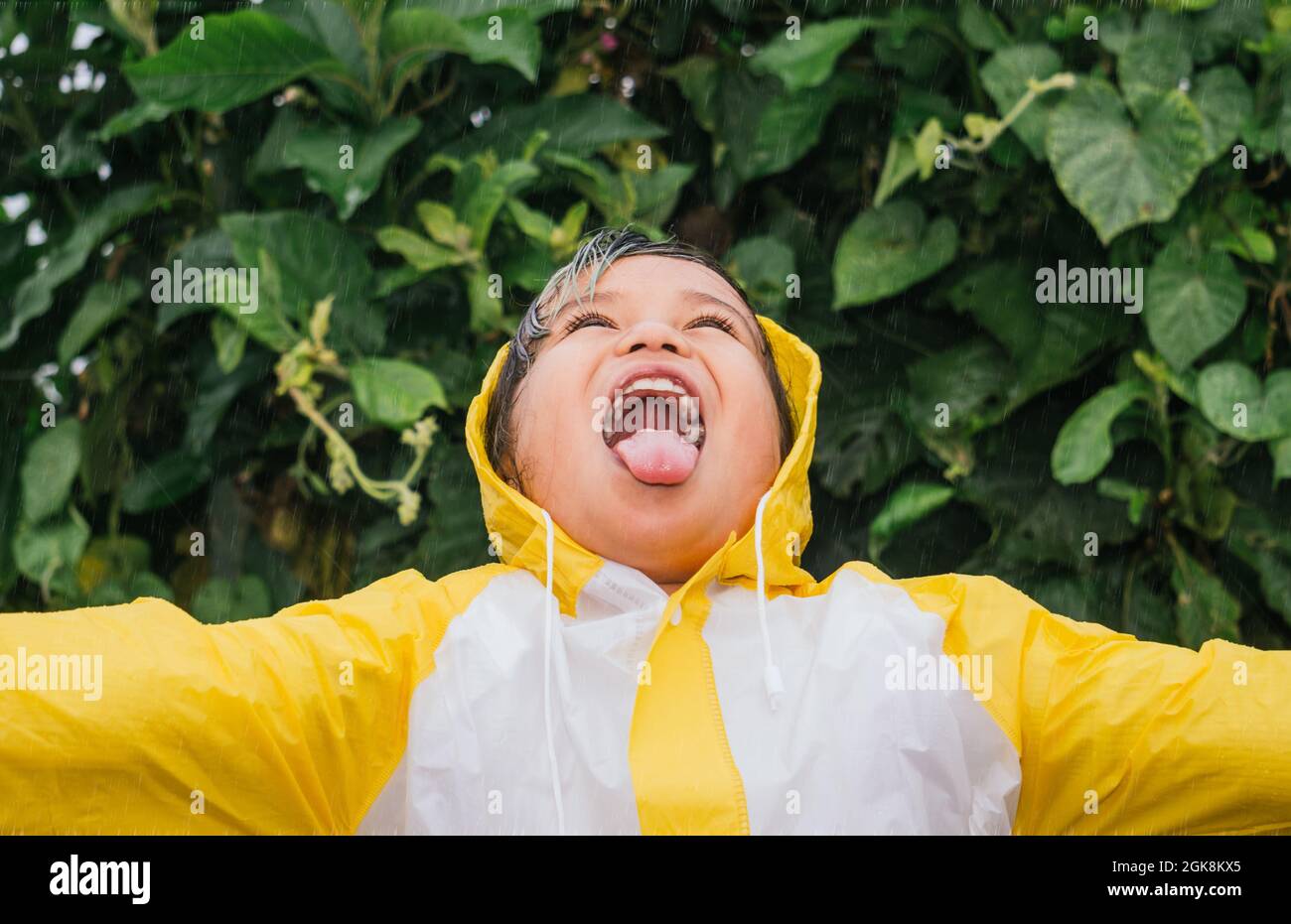 Content ethnic kid in slicker with tongue out catching rain drops while ...
