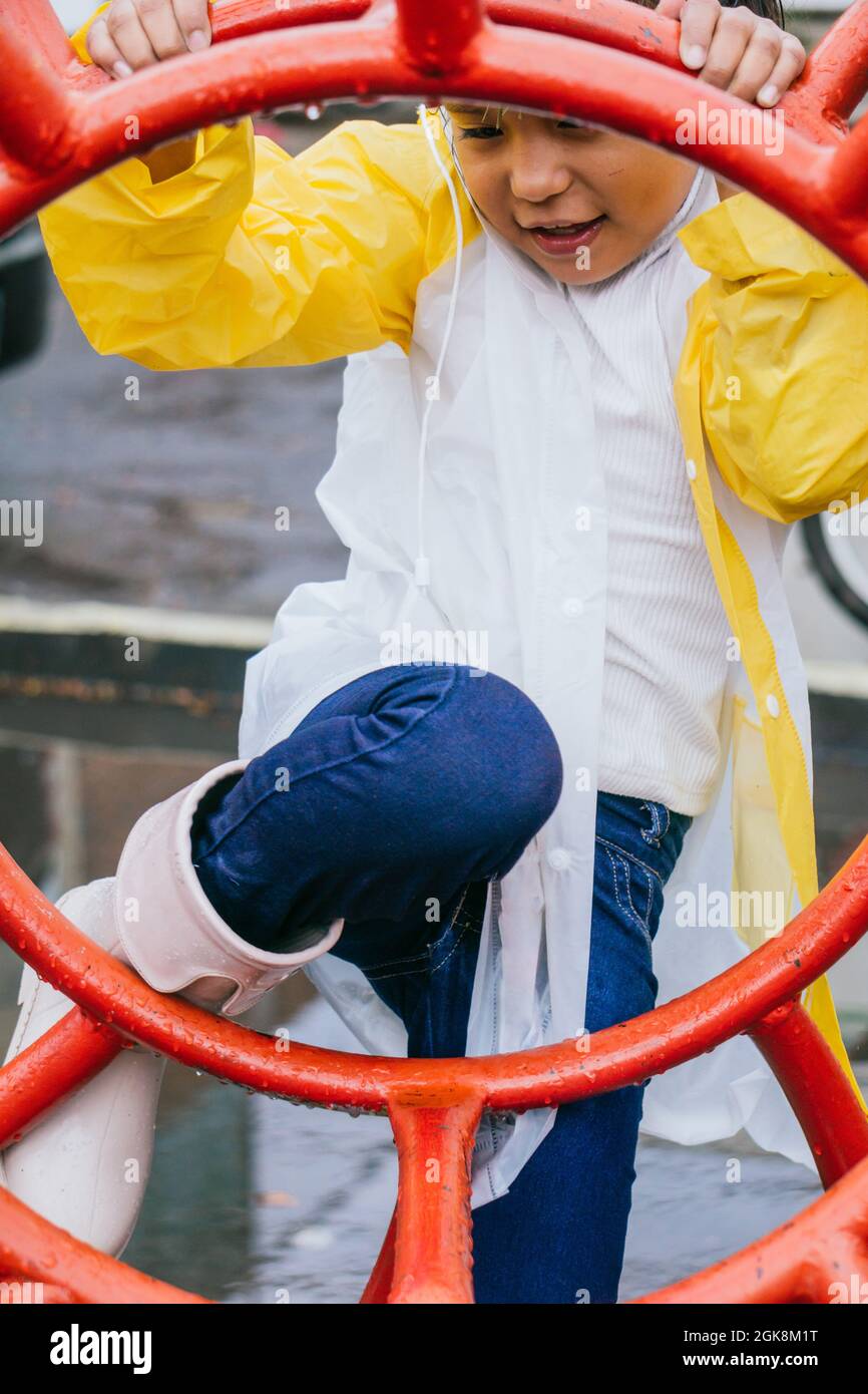 Cheerful ethnic child with wet hair in slicker having fun between metal ...