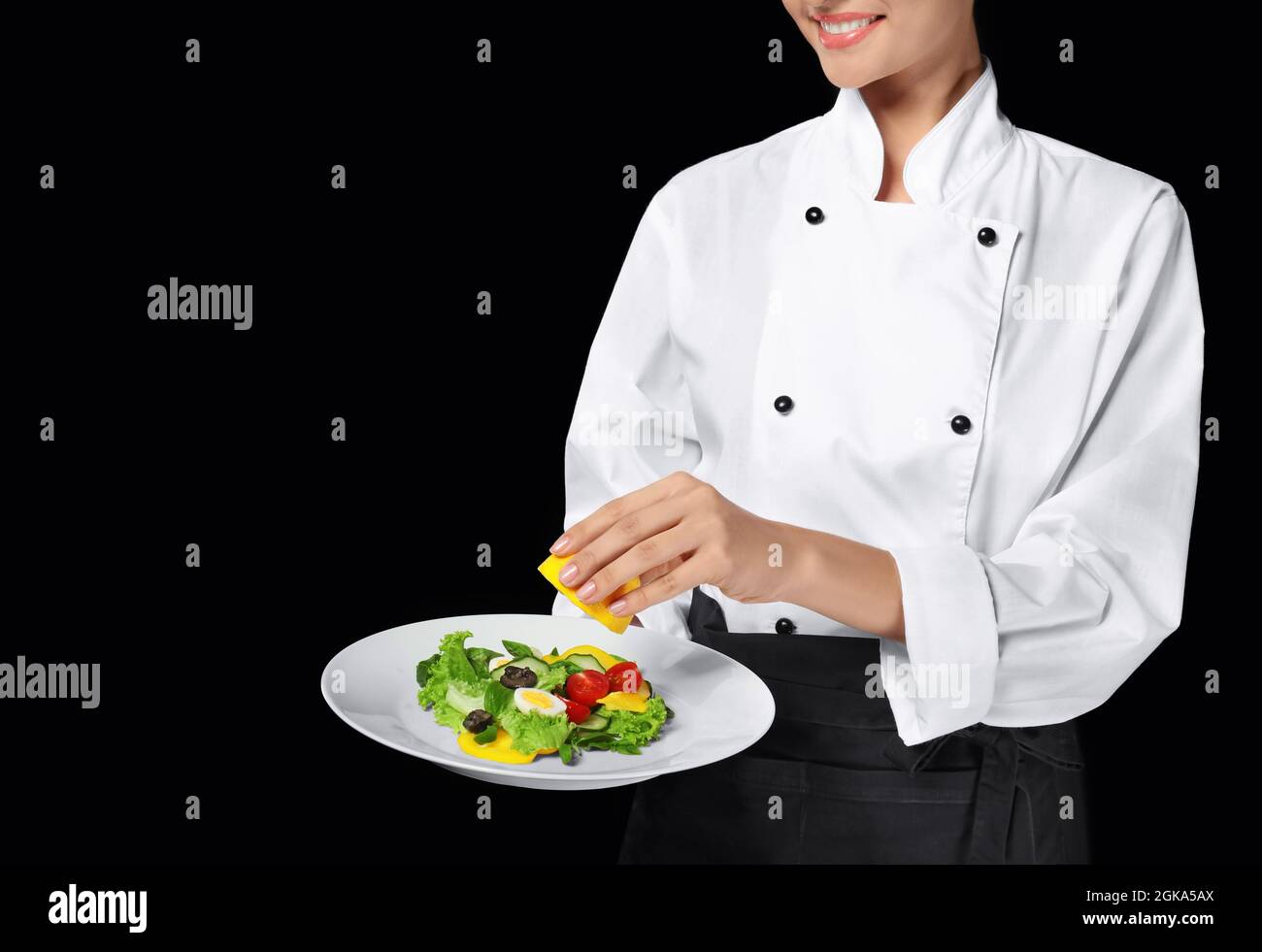 Young female chef dressing salad with lemon juice, on dark background ...
