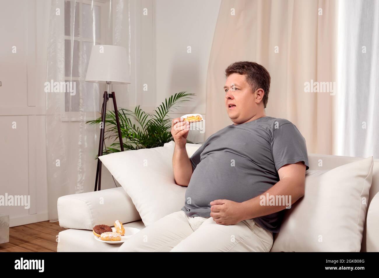 Overweight young man eating sweets on sofa at home Stock Photo - Alamy