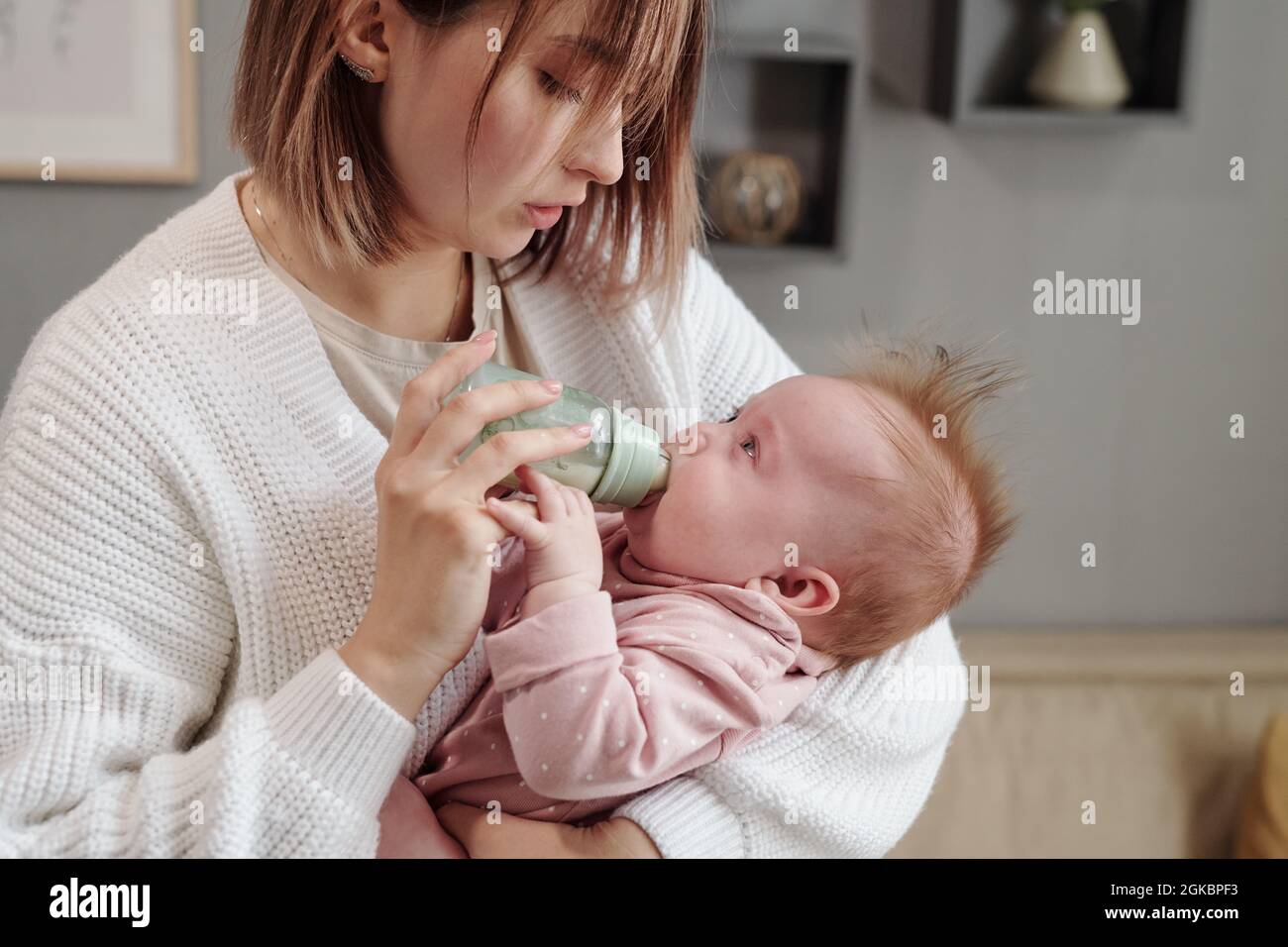 Young careful mother holding cute baby girl and feeding her from bottle ...