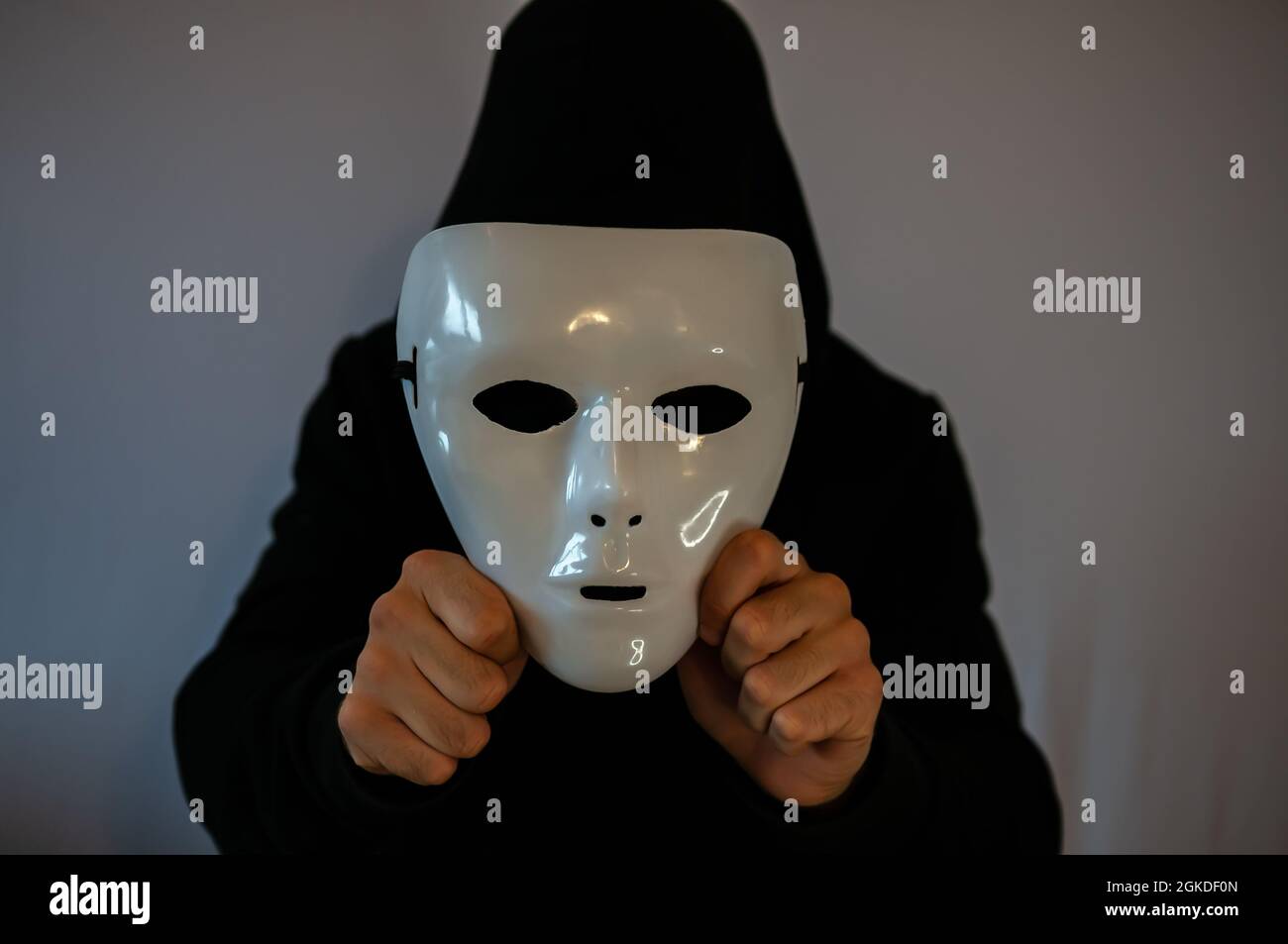 Man with black hood holding an anonymous white mask on dark background ...