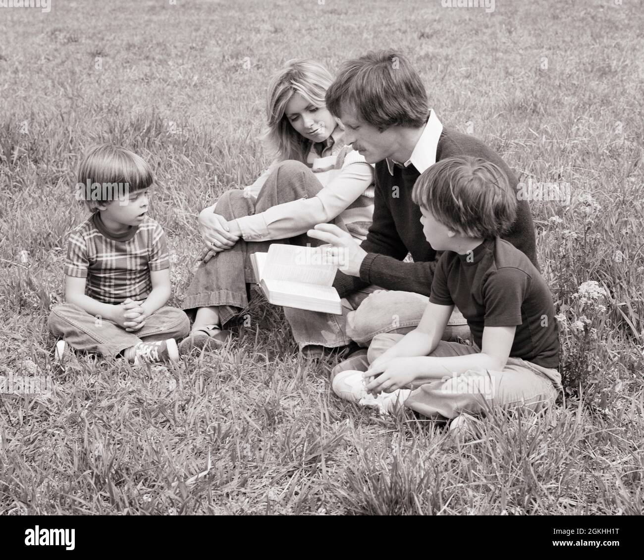 1970s FAMILY MOTHER FATHER TWO SONS SITTING IN GRASS FATHER READING TO ...