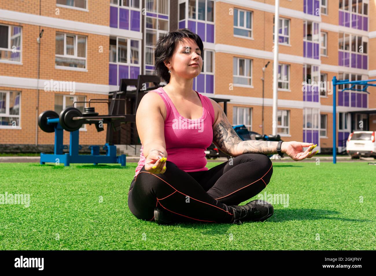 young sturdy chubby woman meditates on the lawn after exercising in the ...
