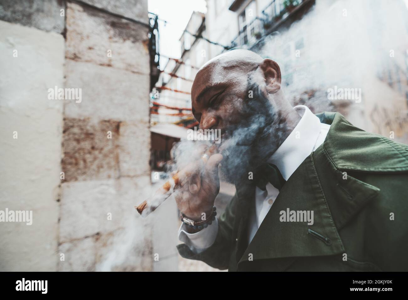 A side view of an elegant mature bearded black guy in a dark-green fashionable outfit smoking a ...