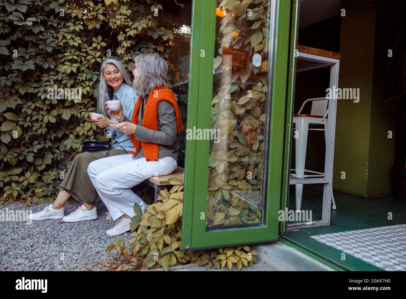 Cheerful senior ladies friends with cups of drinks sit on bench in cafe ...