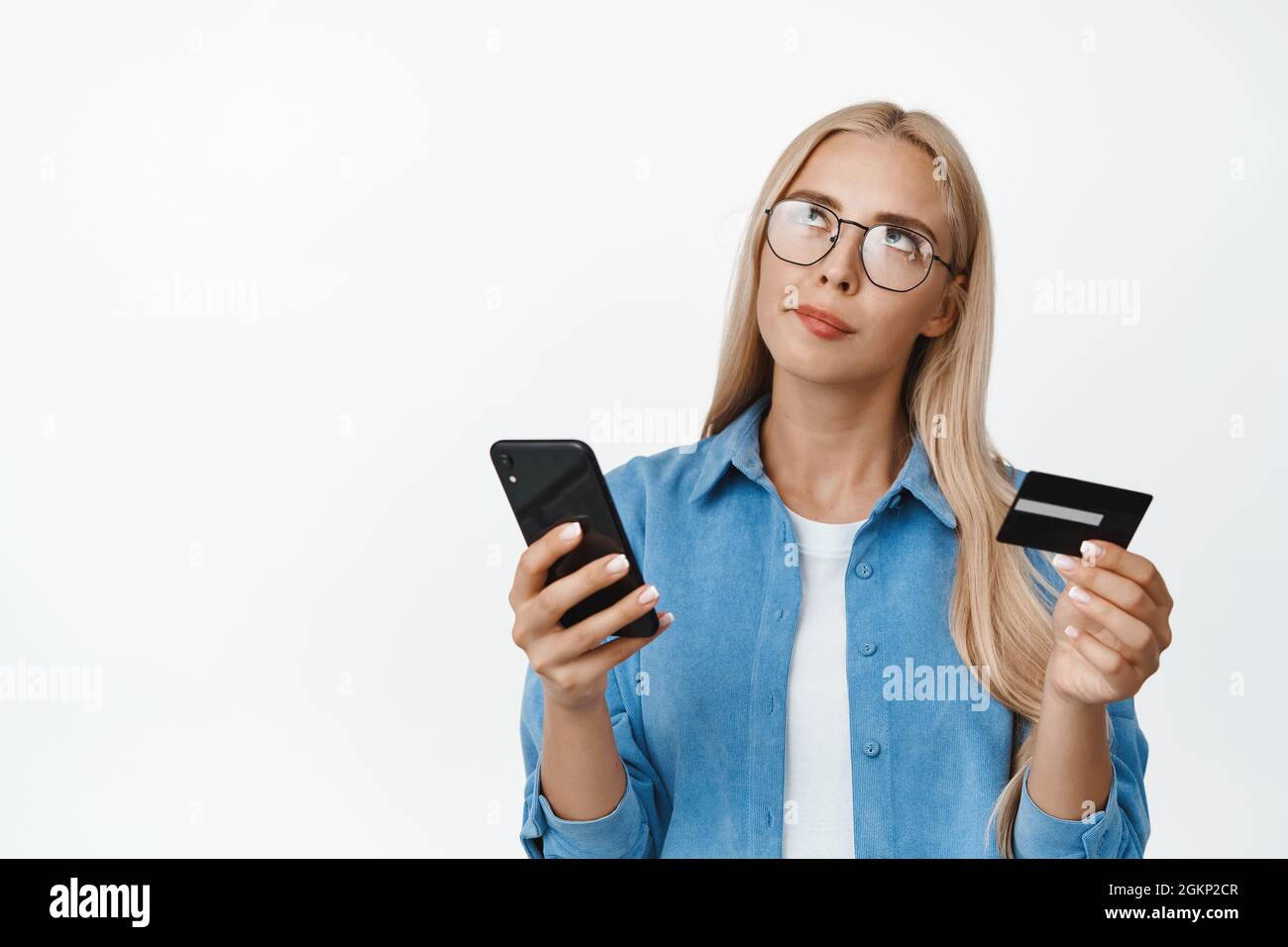Online shopping. Cute young woman thinking, holding smartphone and credit card, using app to buy ...