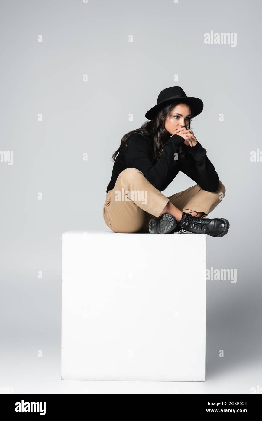 full length of pensive model in fedora hat sitting with crossed legs on ...