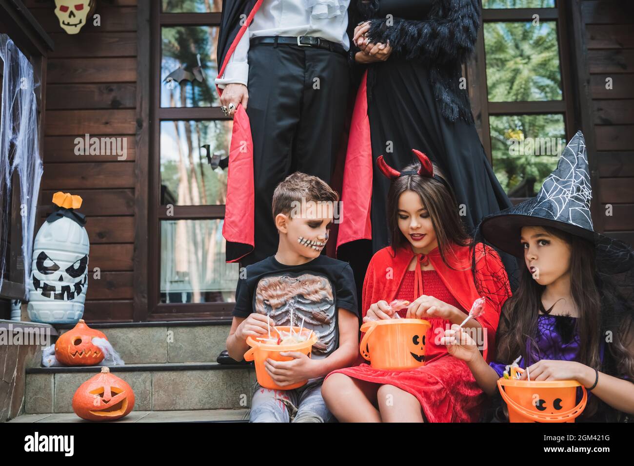 Kids In Halloween Costumes Sitting On Cottage Stairs With Buckets Of Sweets Near Parents Stock kids-in-halloween-costumes-sitting-on-cottage-stairs-with-buckets-of-sweets-near-parents-stock