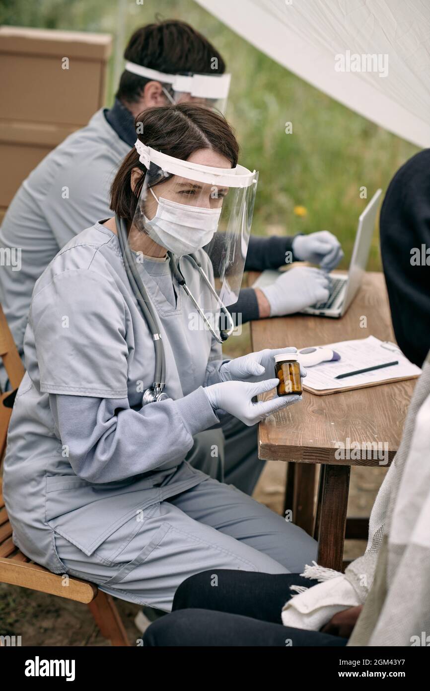 Female nurse in plastic screen mask and gloves sitting at table ...