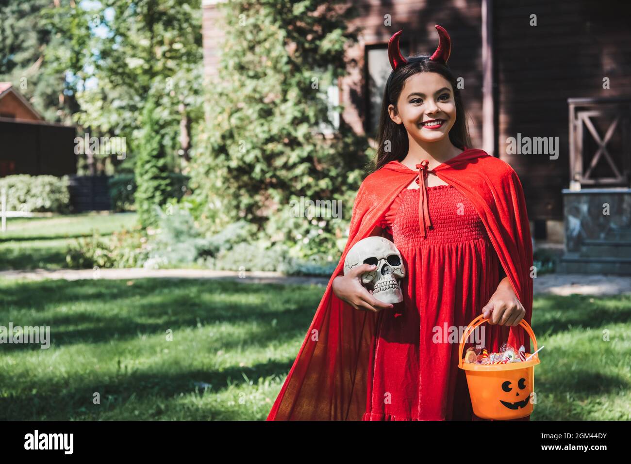 happy child in red demon costume standing with skull and bucket of ...