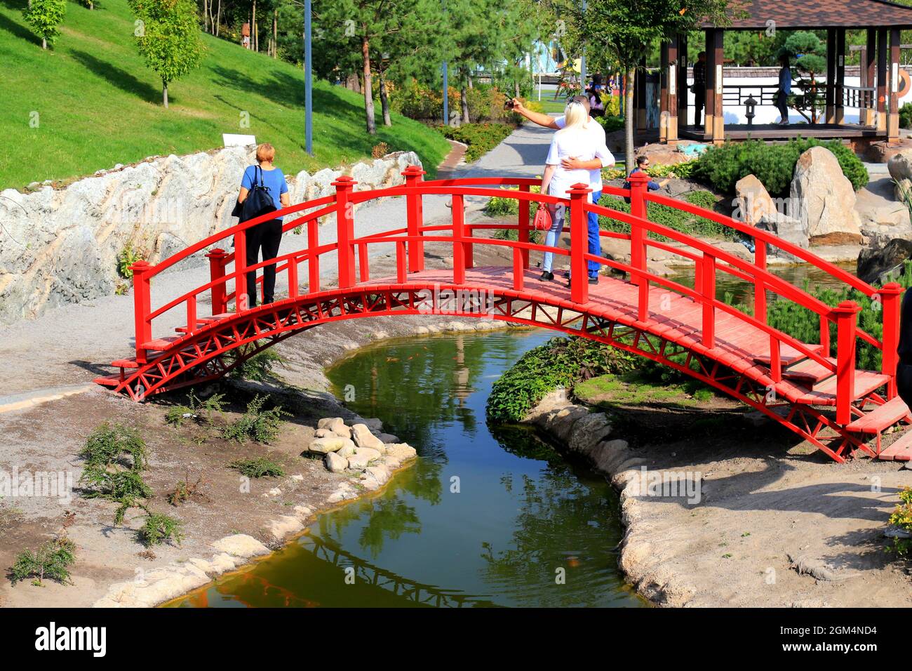 People walk across old red wooden bridge across a small river in a ...