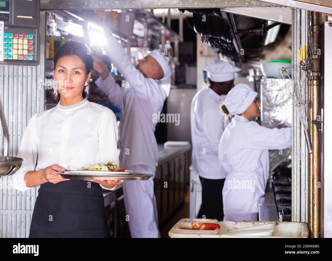positive female waitstaff standing with tray in kitchen in restaurante ...
