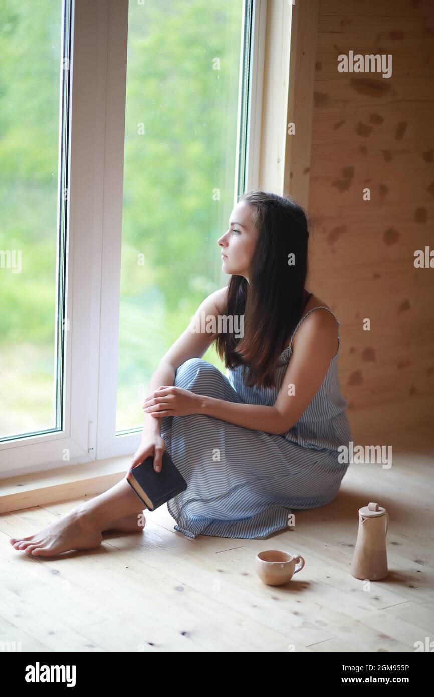 A girl at the window in the house reads a book and drinks tea Stock ...