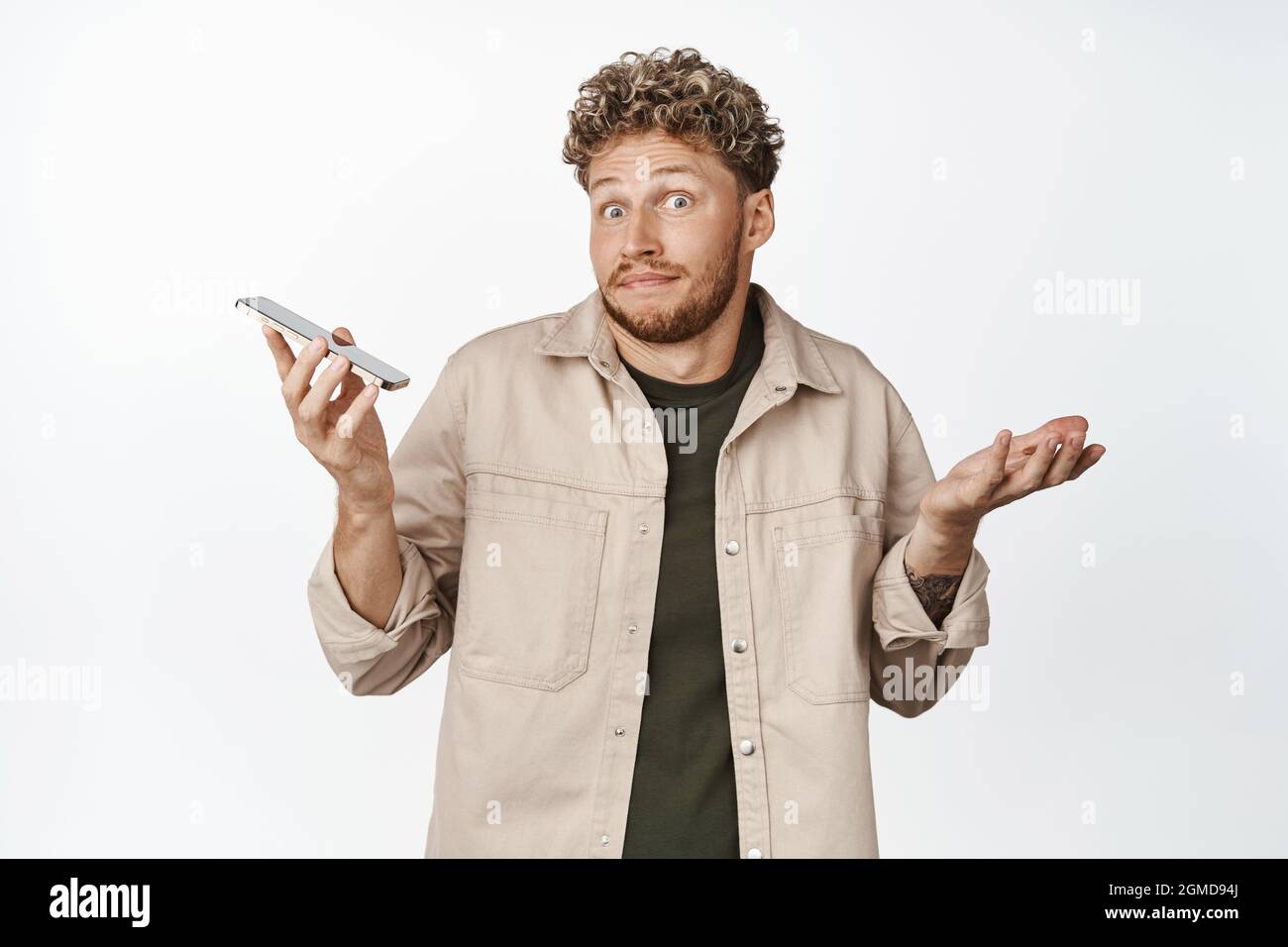 Clueless blond guy shrugging shoulders, holding smartphone and looking ...