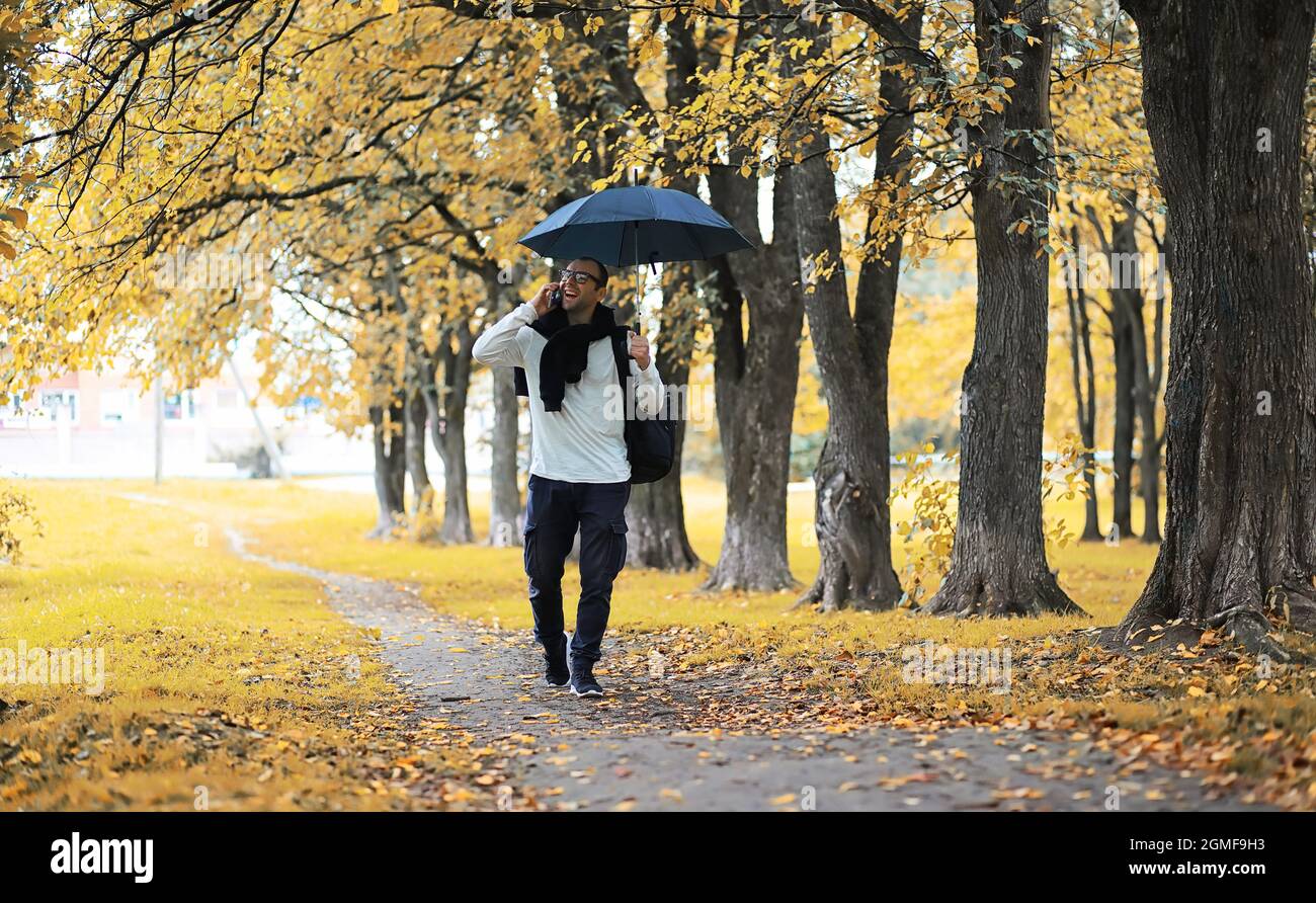 A young man in glasses walks in the park with an umbrella during rain ...