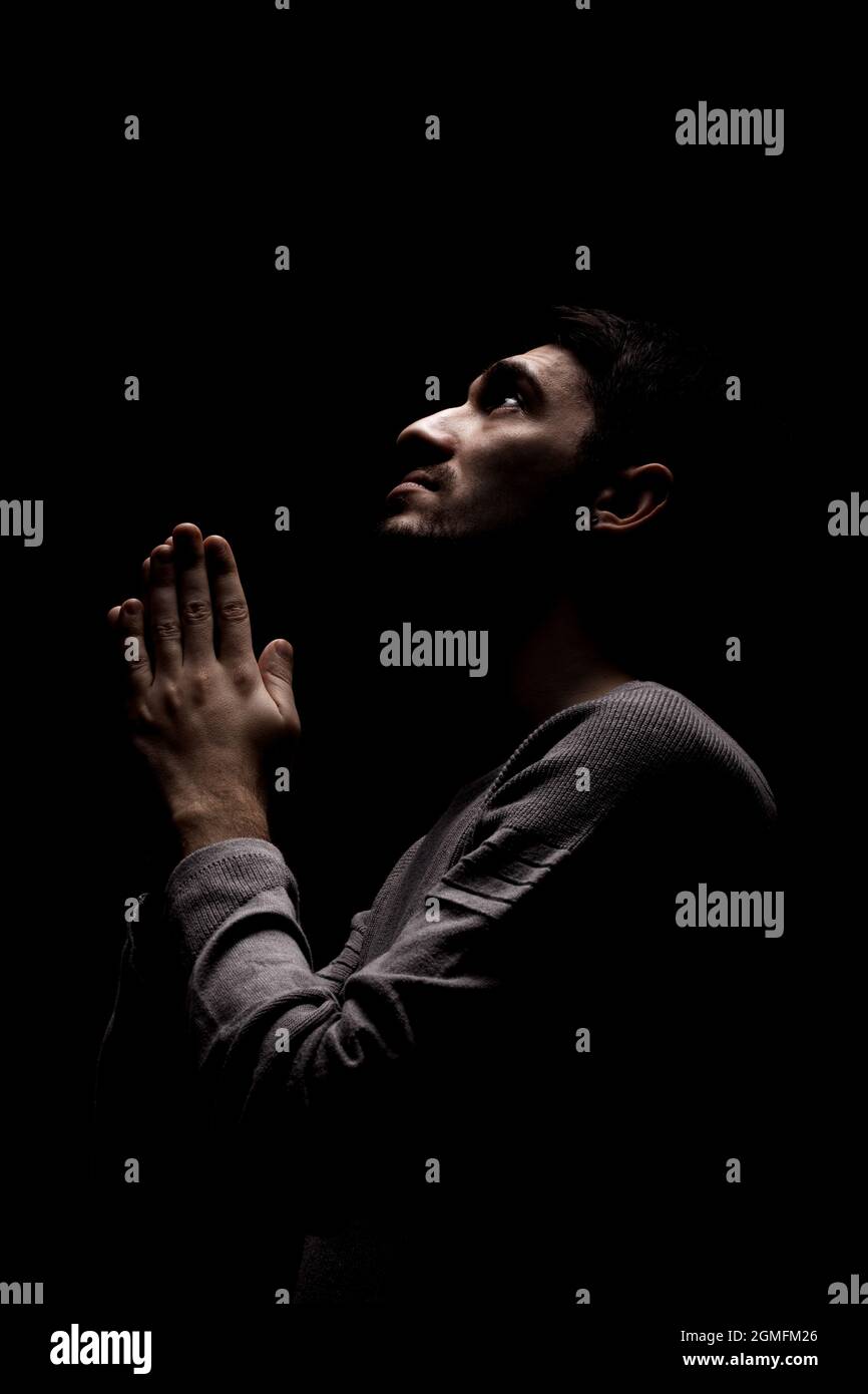 Headshot of serious confident young man praying in dark room. Lit from ...