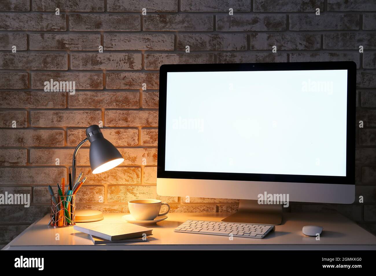 Stylish workplace with glowing lamp and modern computer near brick wall ...